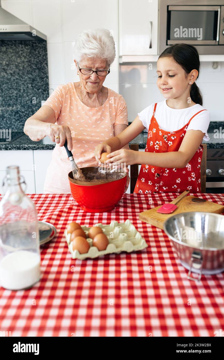 Granny whisking dough near granddaughter breaking fresh egg into bowl while cooking in kitchen ...