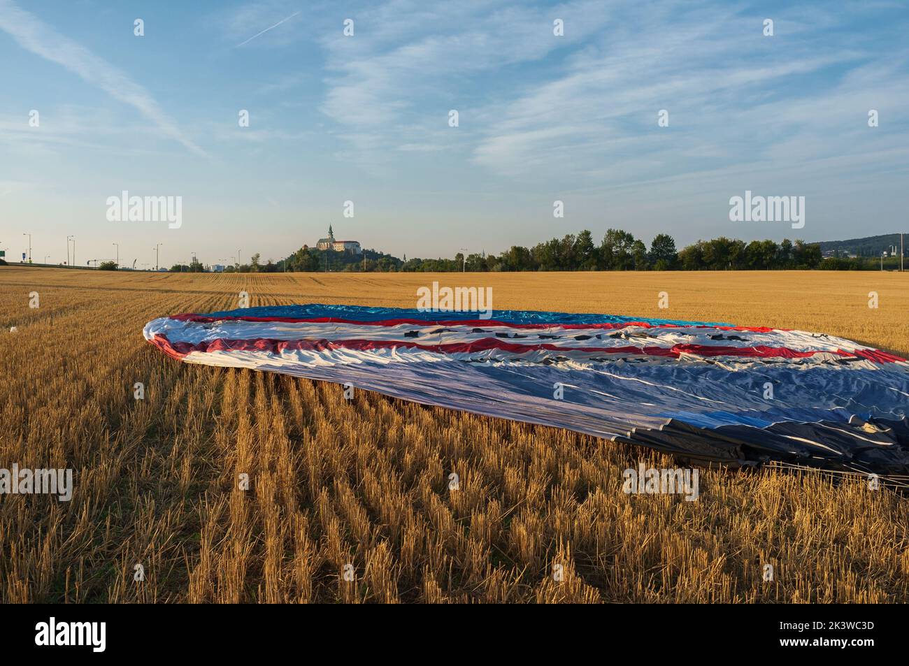 A yellow fild view with an empty flat hot air balloon on green trees ...