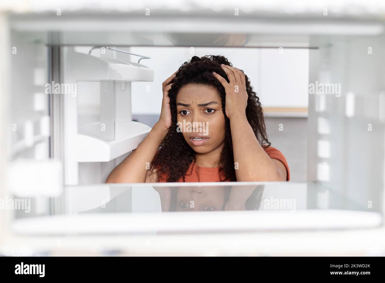 Sad shocked hungry millennial black lady hold her head, looking into empty refrigerator ...