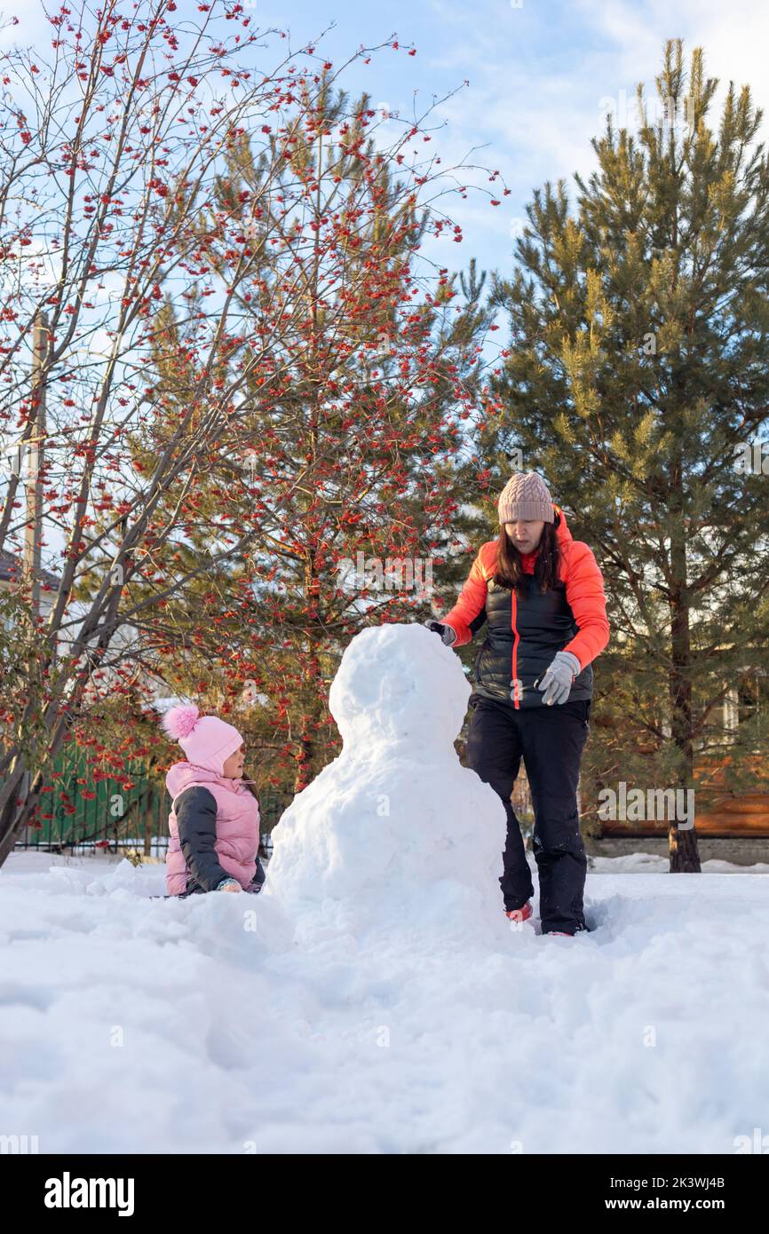 Portrait of little girl sitting on snow and mother making snowman on ...