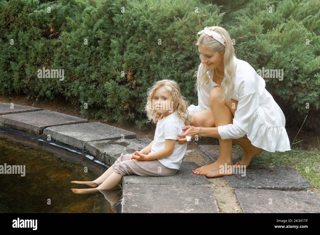 Portrait of little girl sitting on edge of pond with feet in water in ...