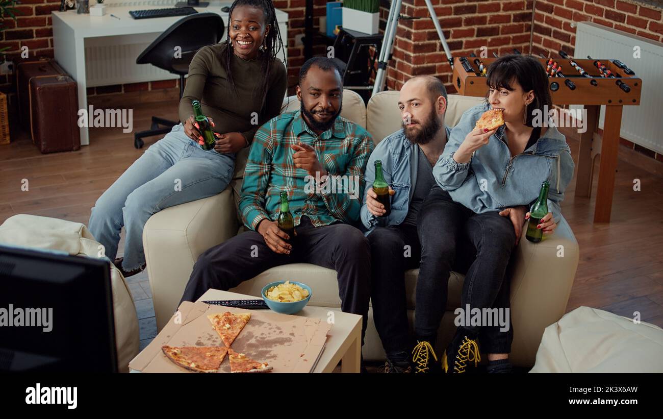 Multiethnic group of friends clinking beer bottles at house party, making toast and watching ...