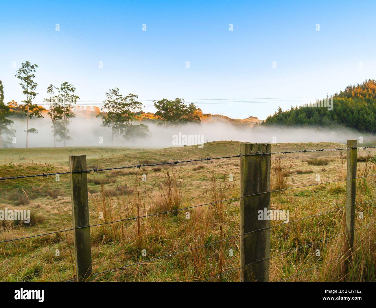Mist in valley in morning light beyond fence and fields under blue sky ...