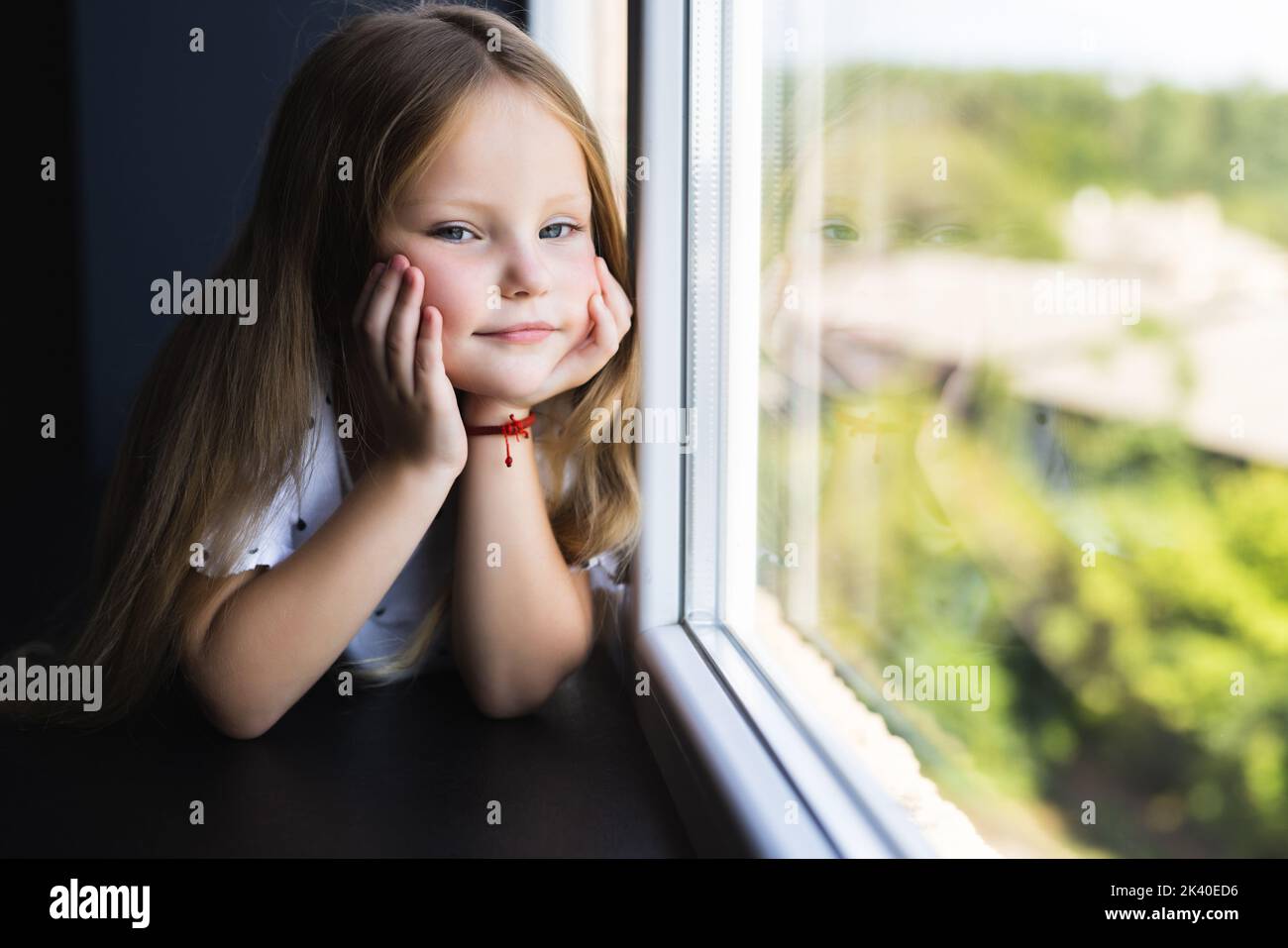 Beautiful little girl smiling and watching out the window. A child looks out the window. Young ...
