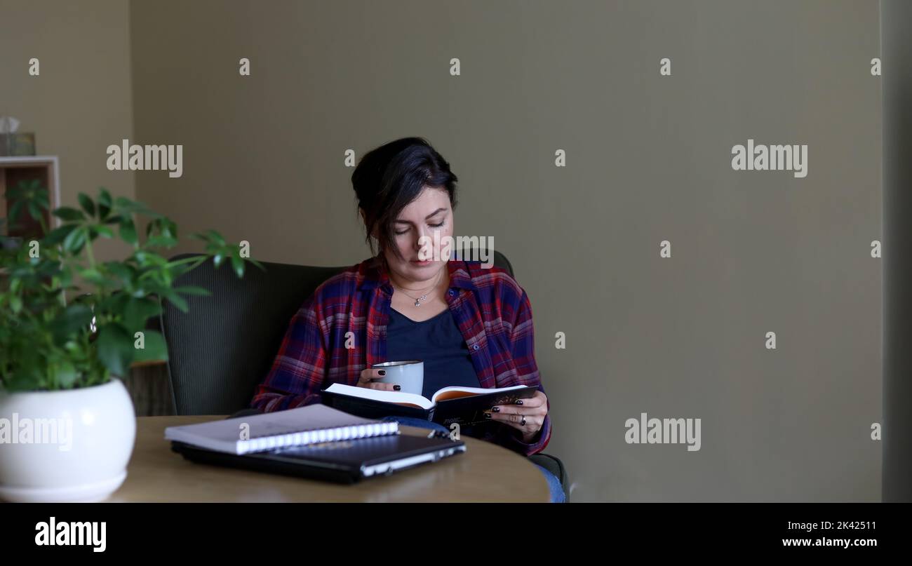 Brunette woman sit with book and cup of tea in armchair at table in ...