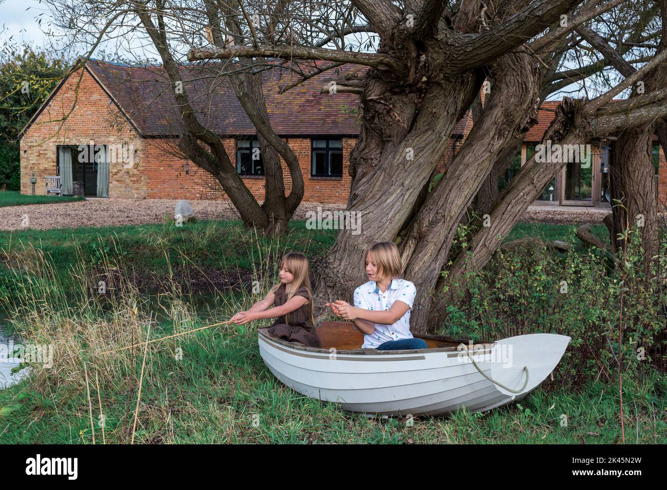 Boy and girl playing in fishing boat on land, fishing in pond by house ...