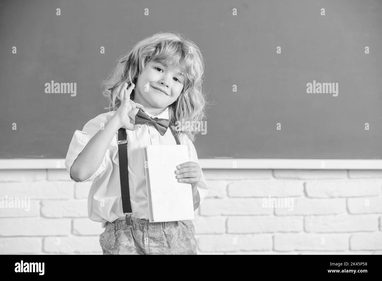 cheerful kid boy in bow tie in school classrrom with book show ok ...
