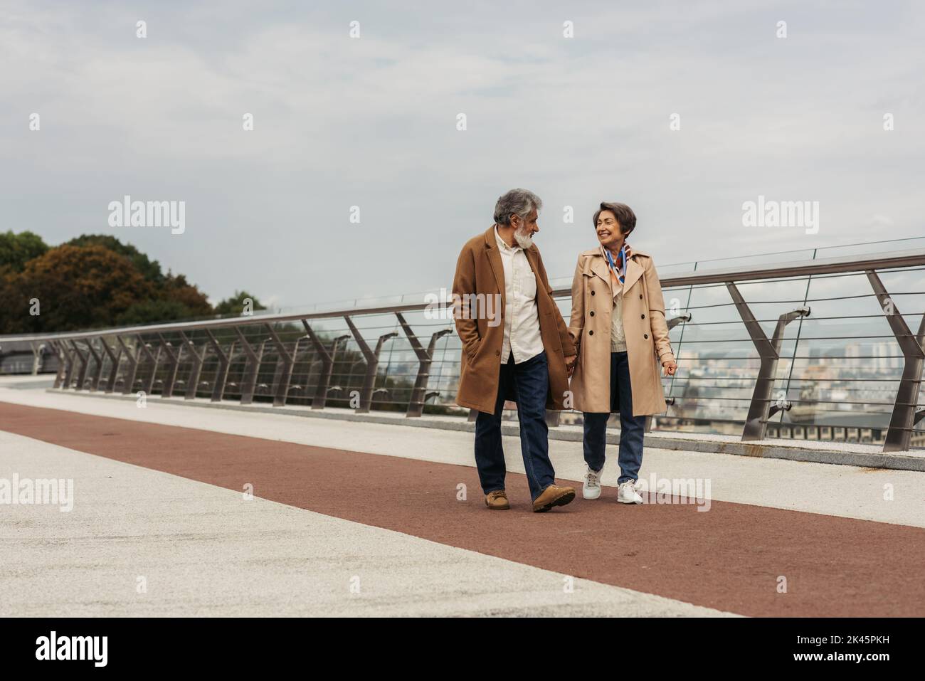 full length of pleased senior couple in beige coats holding hands and ...