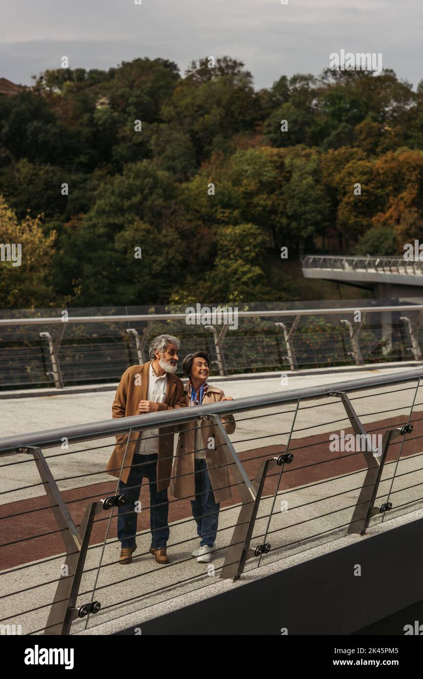 full length of happy senior woman and bearded man standing near bridge ...