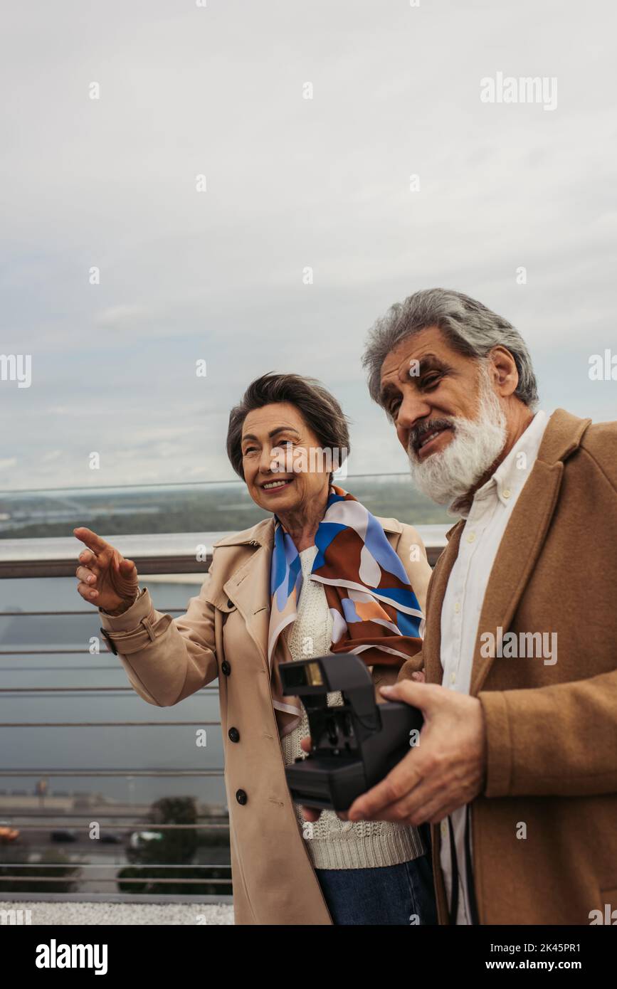 happy senior man holding vintage camera near wife smiling and pointing ...