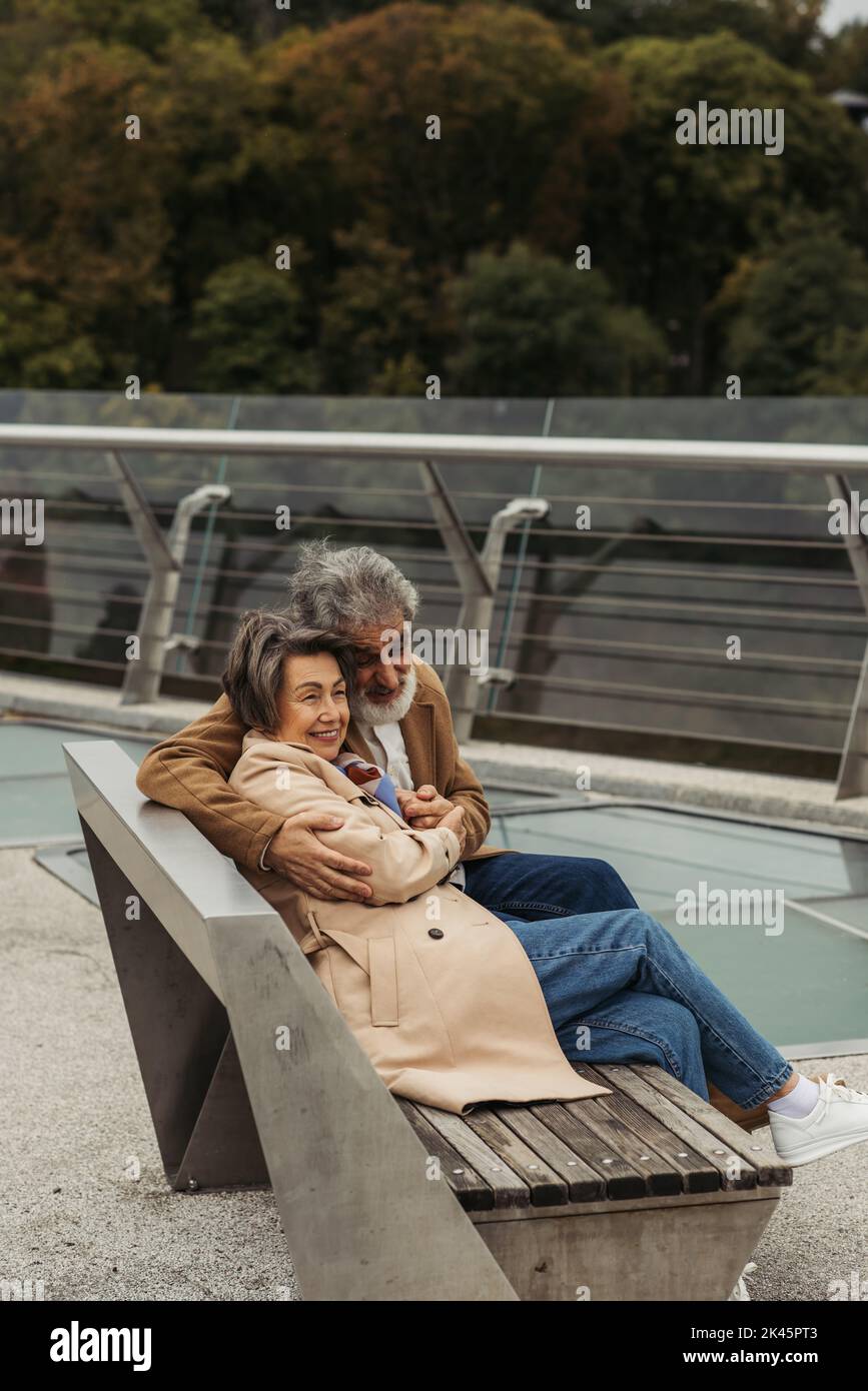 happy senior man hugging elderly wife while sitting on bench near ...