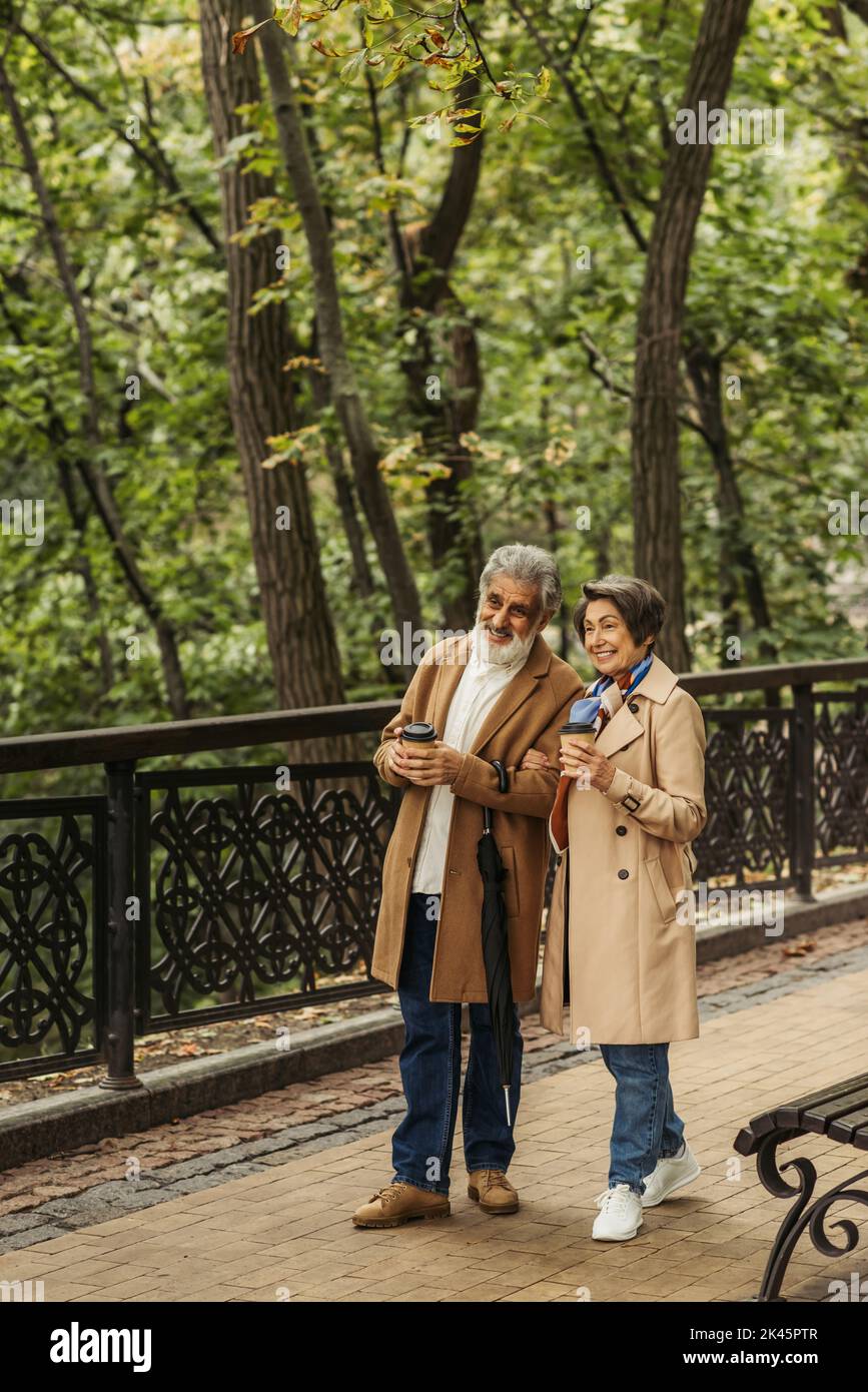 full length of cheerful senior couple in beige coats holding paper cups ...