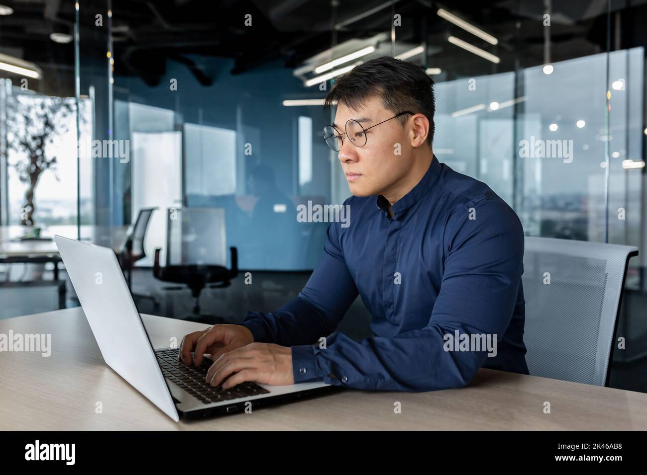 Serious and focused businessman working with laptop, man in glasses and shirt inside office ...