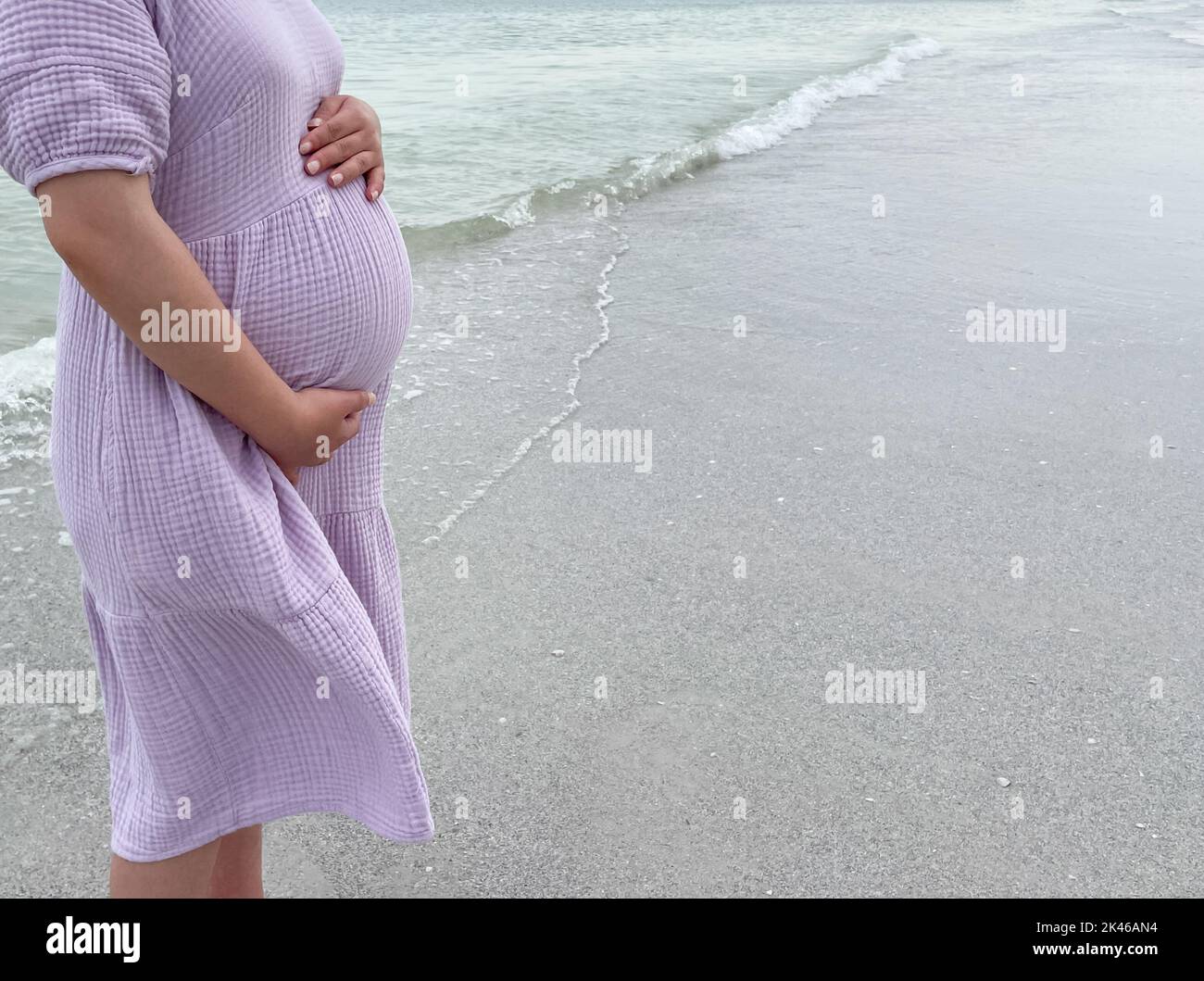 Shot of a pregnant woman standing on the beach with her hand holding ...