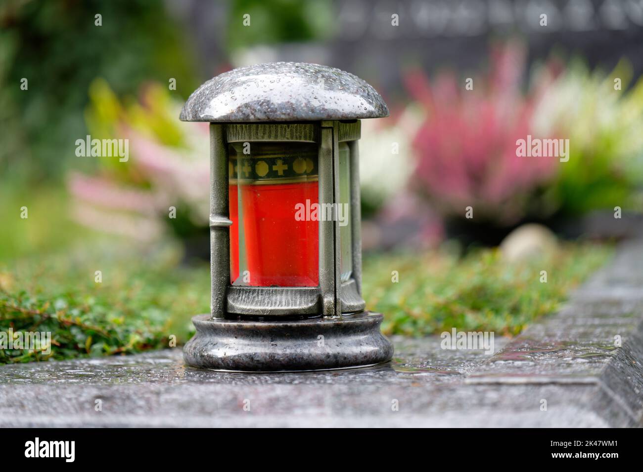 grave lantern with red memorial candle on a grave in front of flower decoration in blurred ...