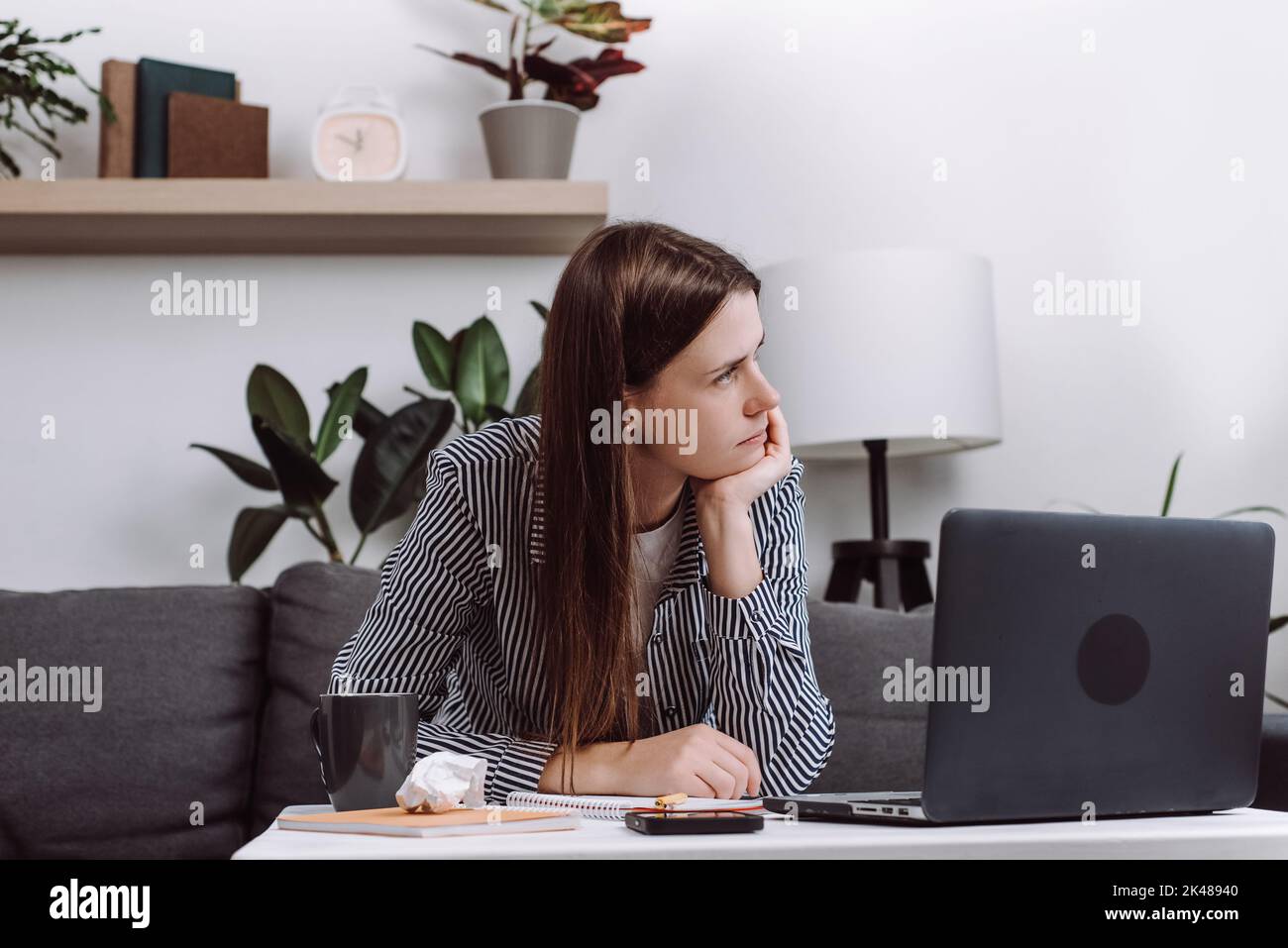 Thoughtful young woman sitting on sofa with laptop thinking of ...