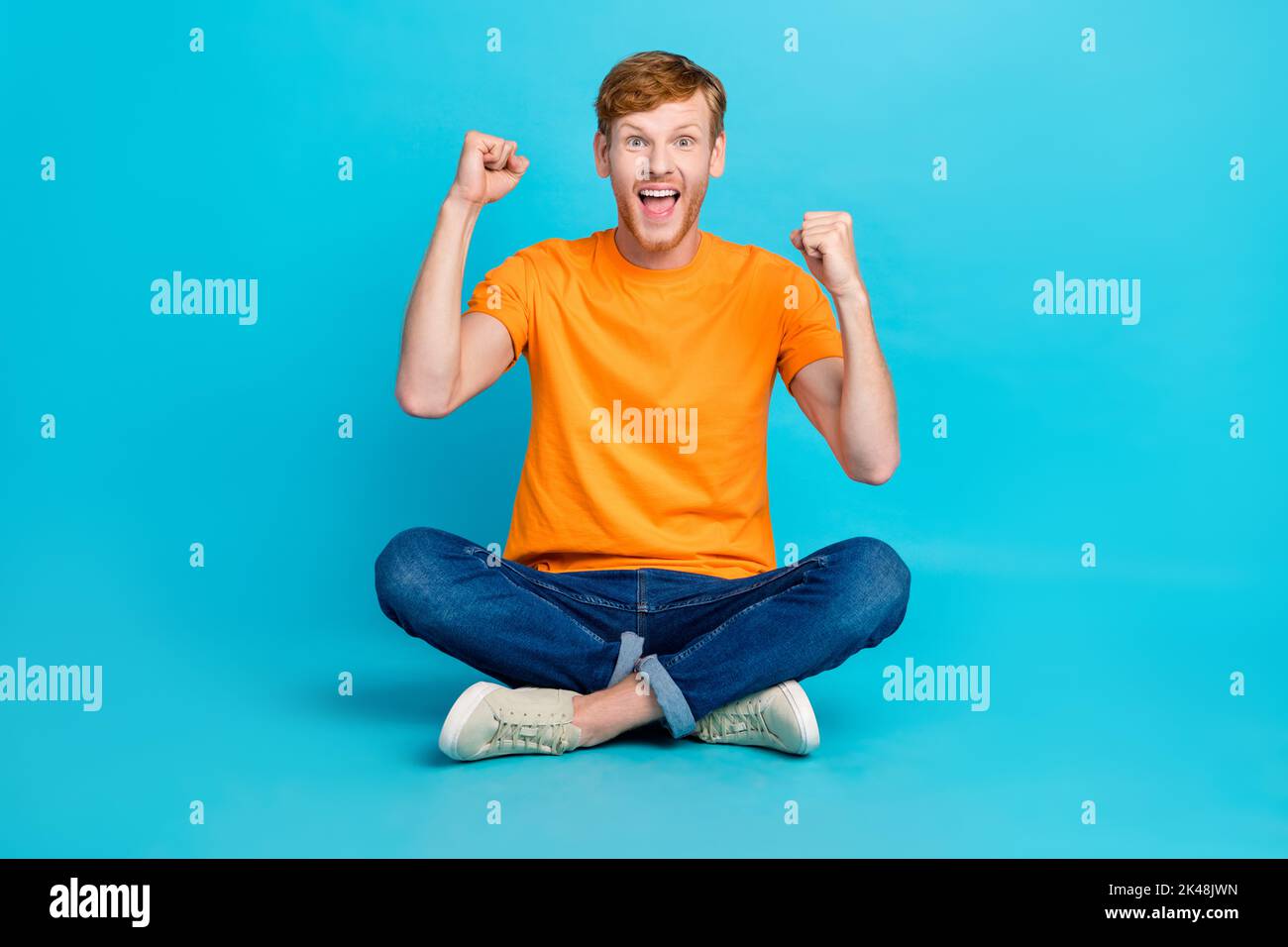 Full Length Photo Of Attractive Lucky Man Wear Orange T Shirt Sitting Floor Rising Fists
