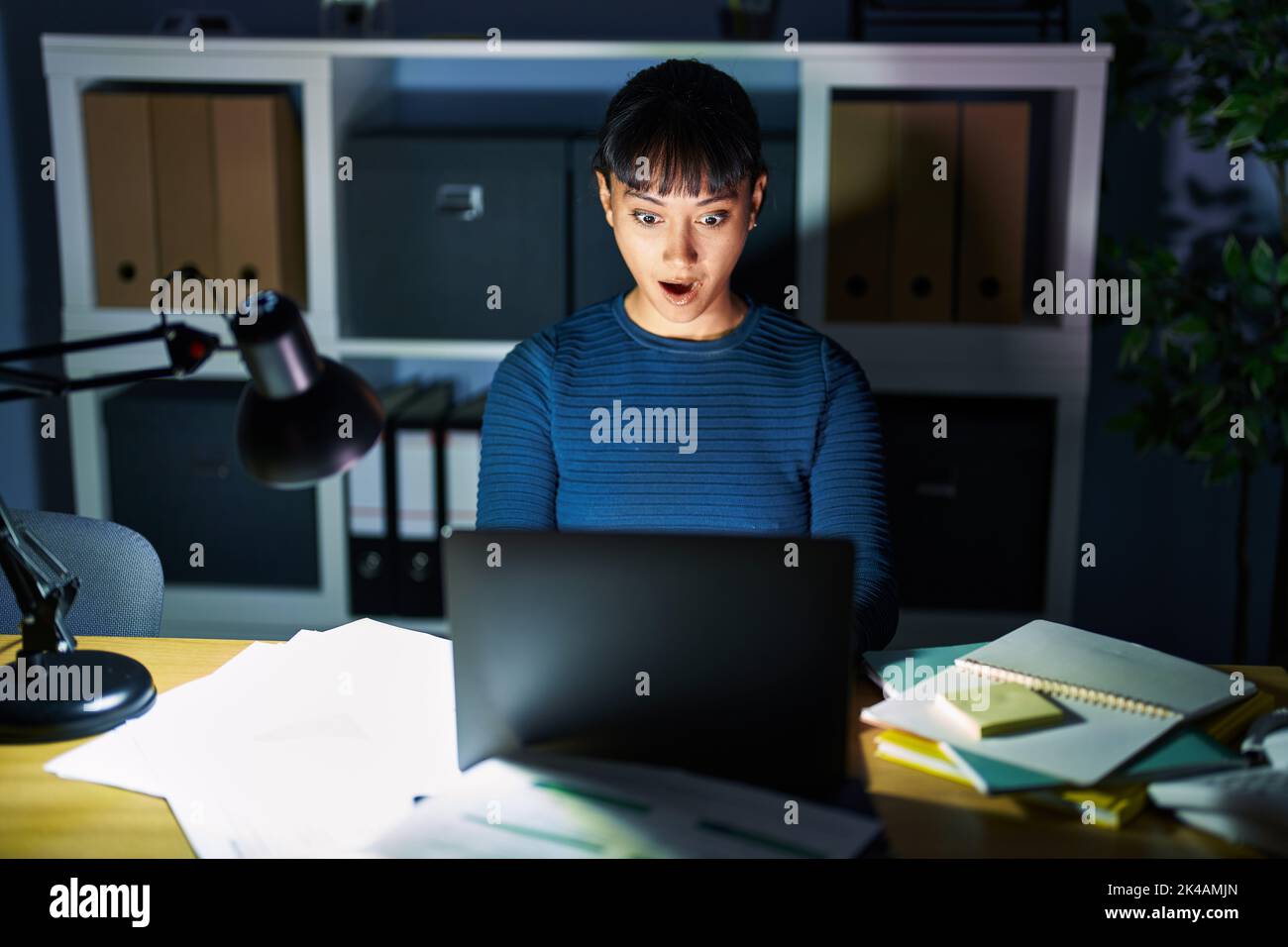 Young beautiful woman working at the office at night afraid and shocked ...