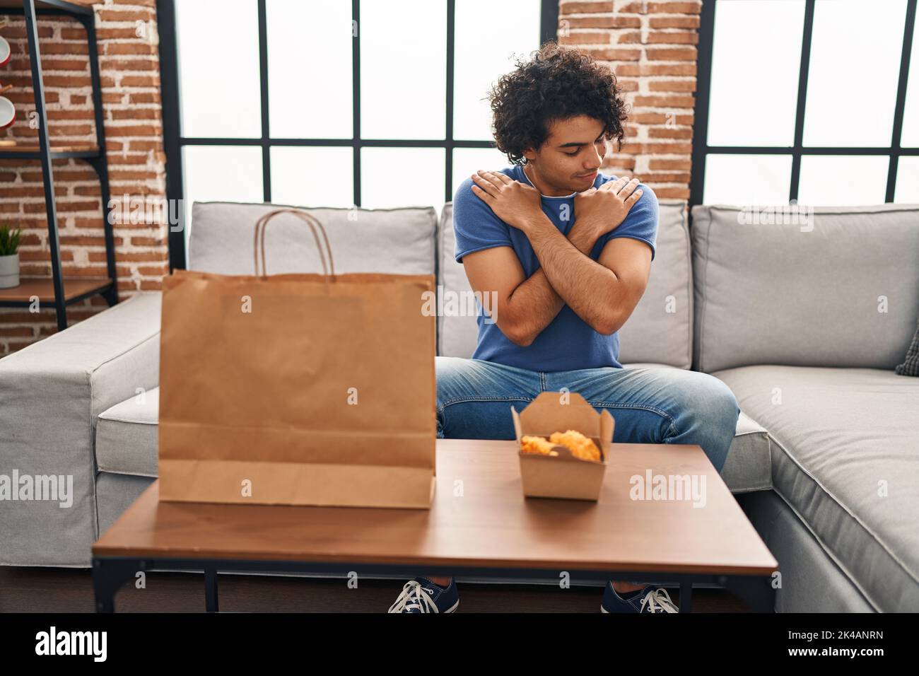Hispanic man with curly hair eating chicken wings hugging oneself happy ...