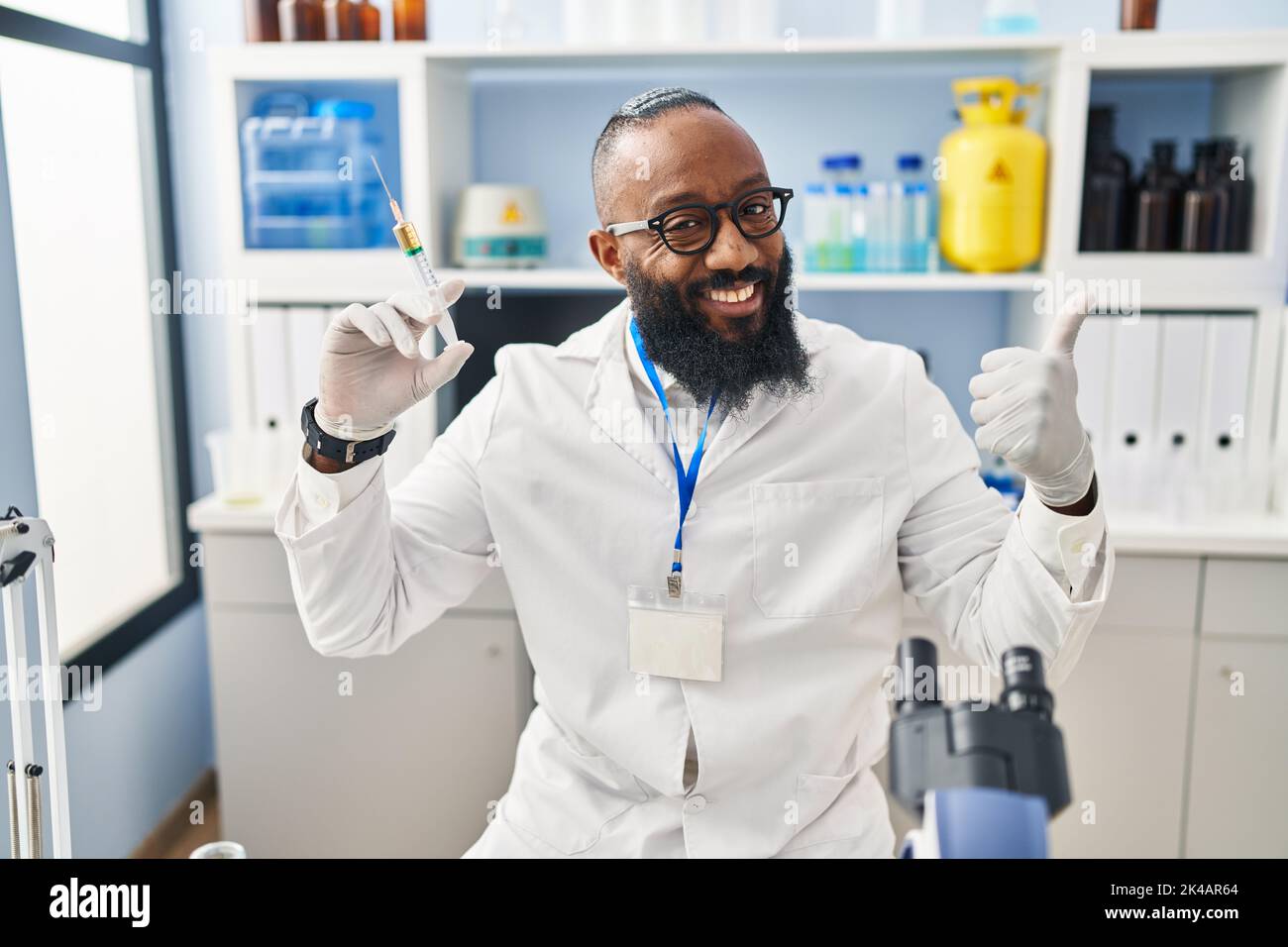 African american man working at scientist laboratory holding syringe smiling happy and positive ...
