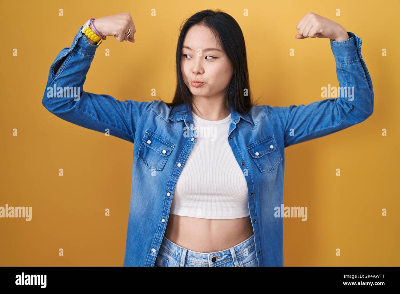Young asian woman standing over yellow background showing arms muscles ...