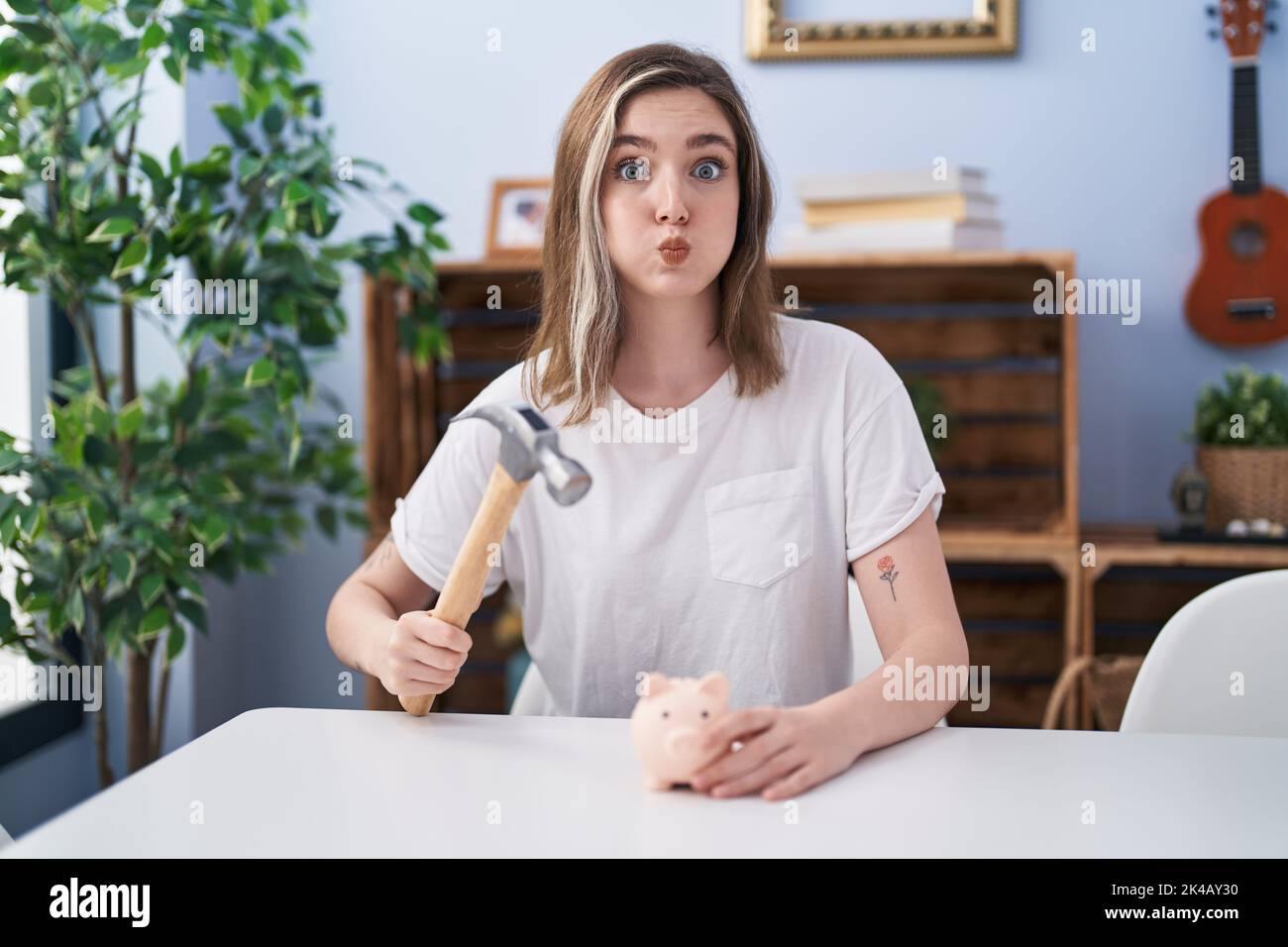 Blonde caucasian woman holding piggy bank and hammer puffing cheeks ...