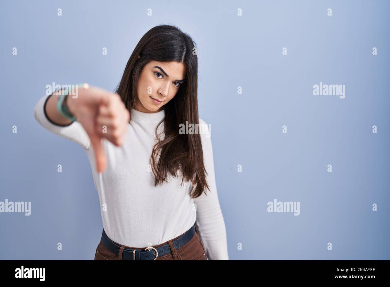 Young brunette woman standing over blue background looking unhappy and ...