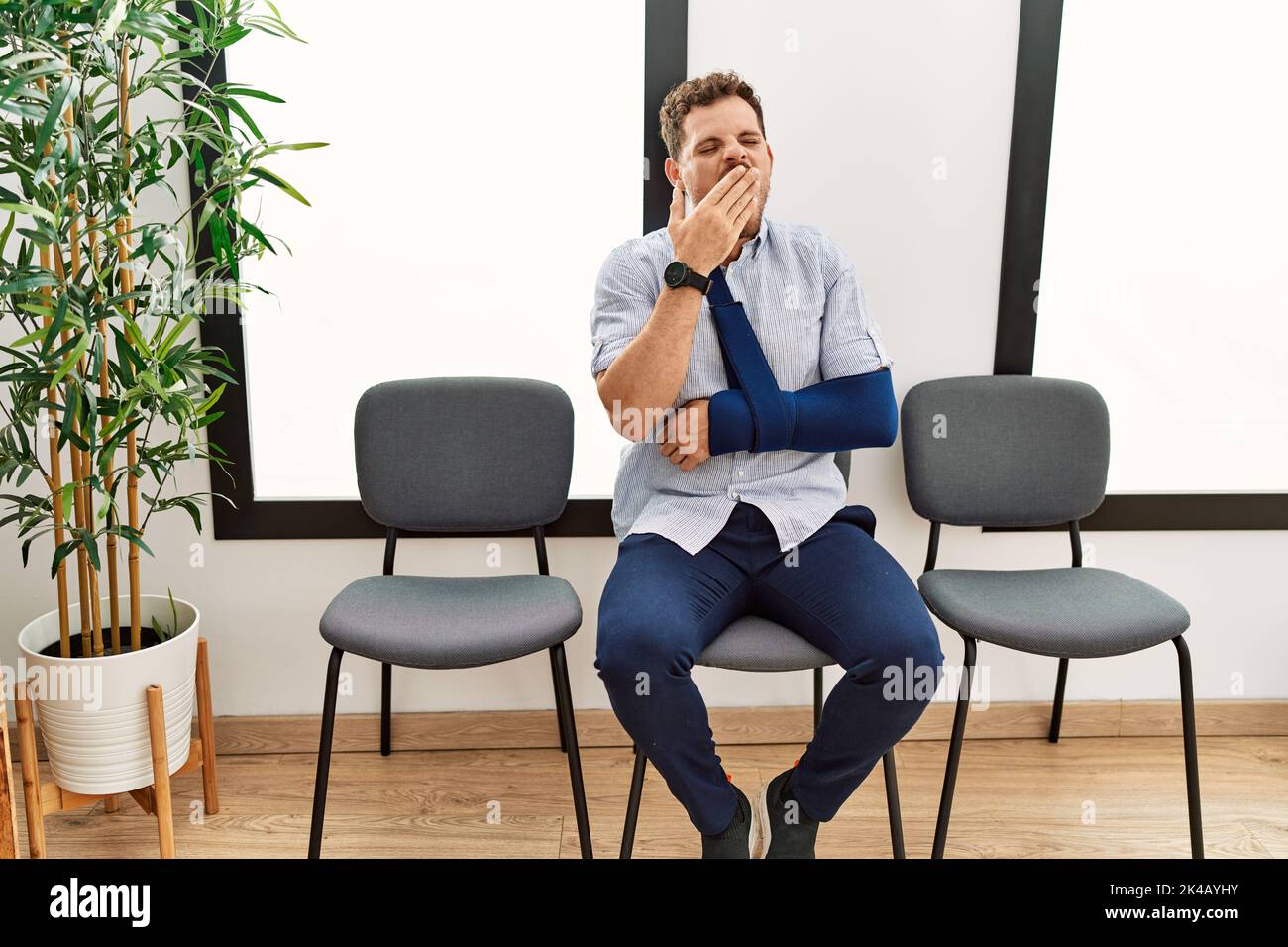Handsome young man sitting at doctor waiting room with arm injury bored ...