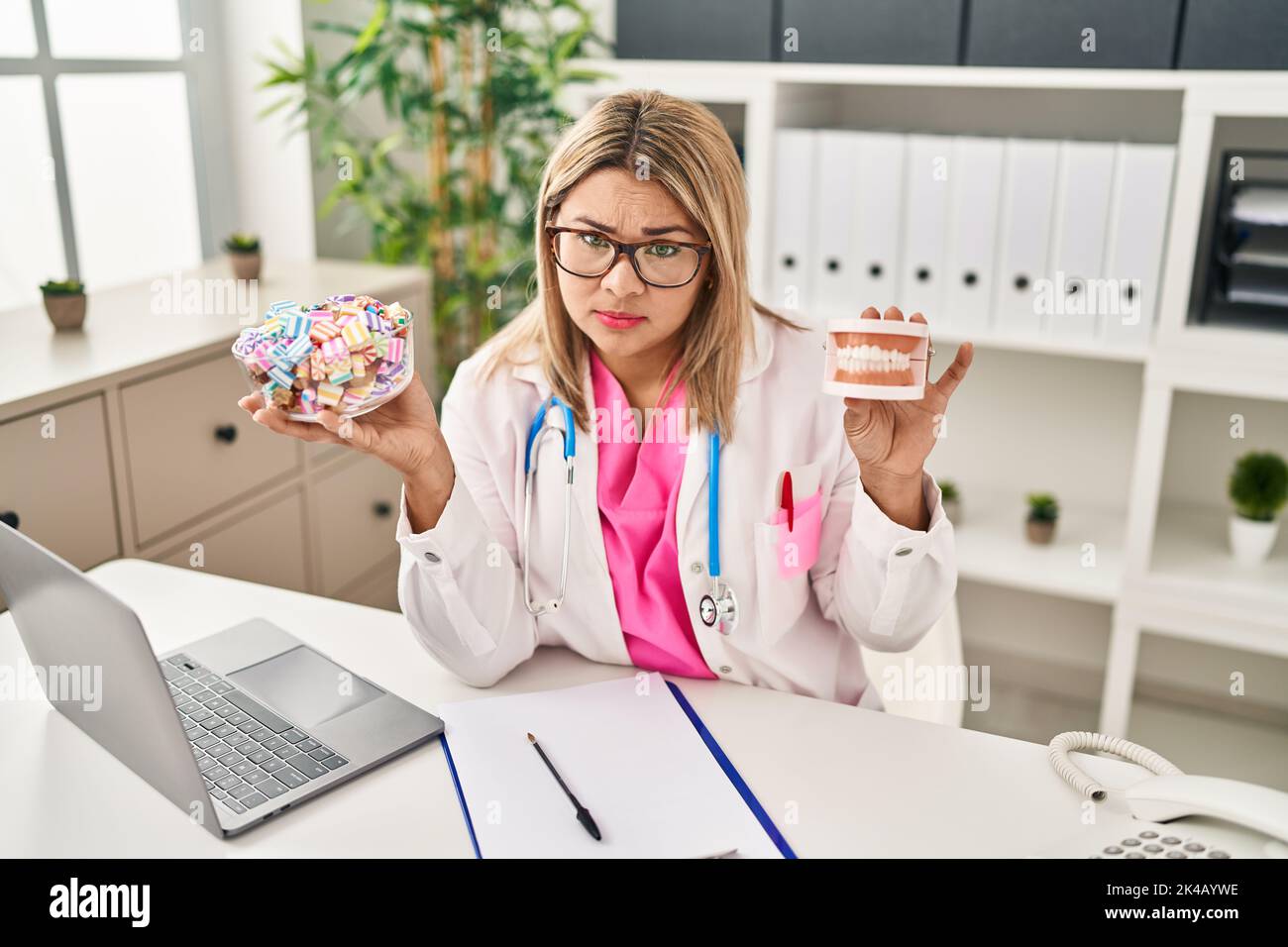 Young hispanic dentist woman holding denture and sweets depressed and ...