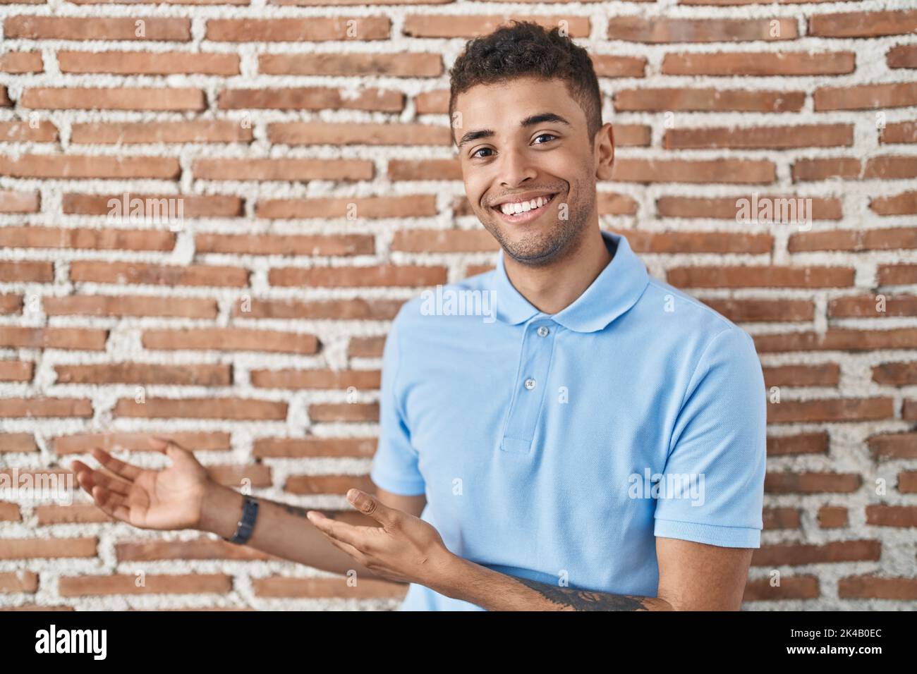 Brazilian young man standing over brick wall inviting to enter smiling ...