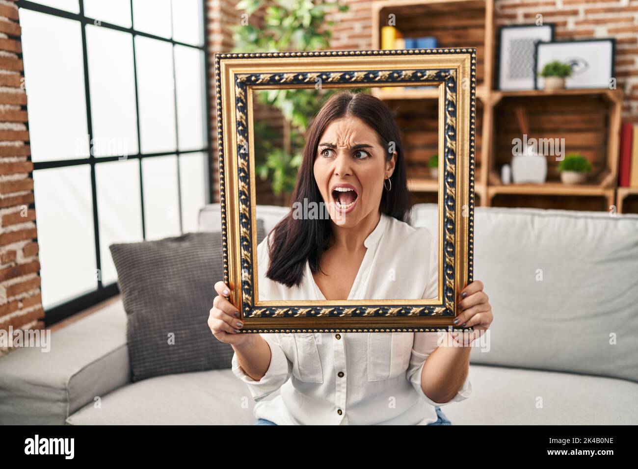 Young hispanic woman holding empty frame angry and mad screaming ...