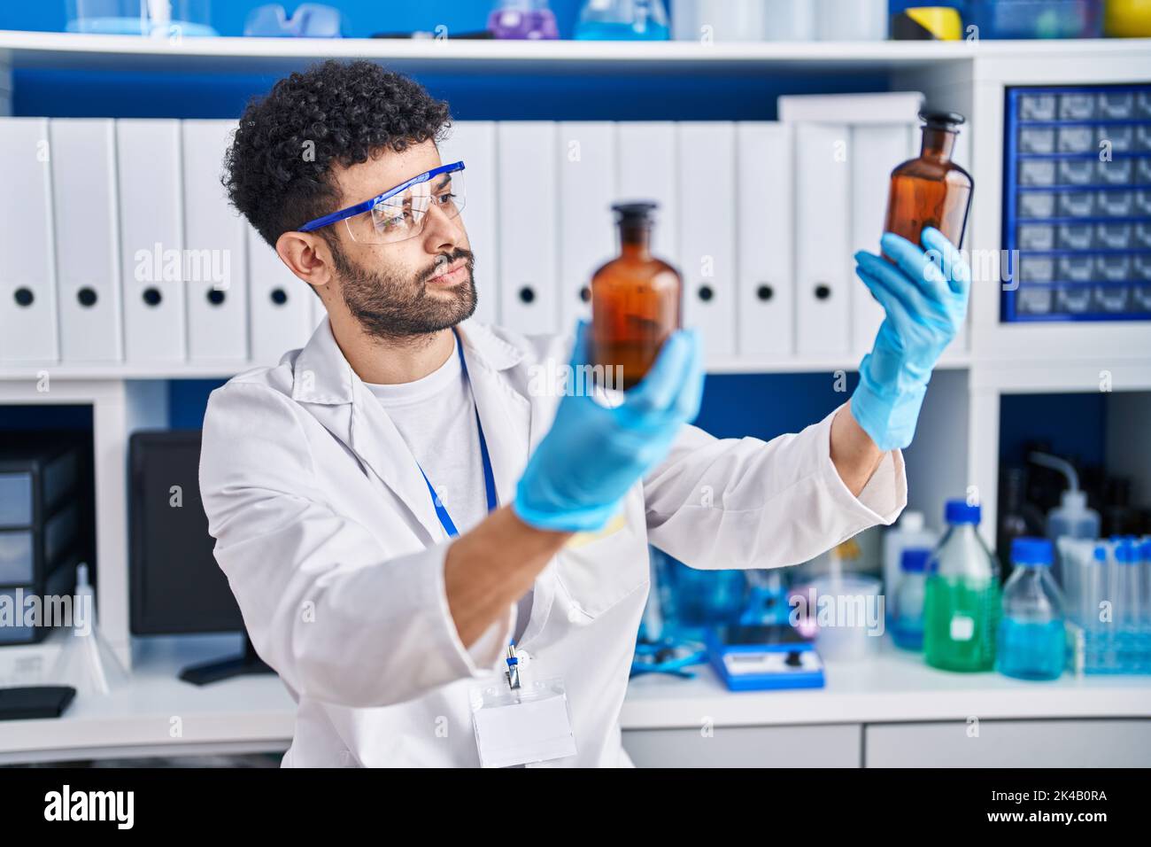 Young arab man wearing scientist uniform holding bottles at laboratory ...