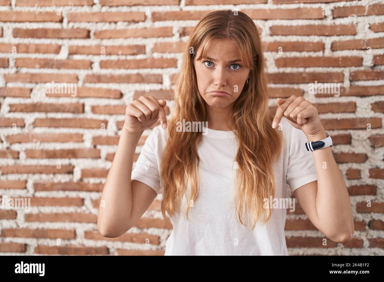 Young caucasian woman standing over bricks wall pointing down looking ...