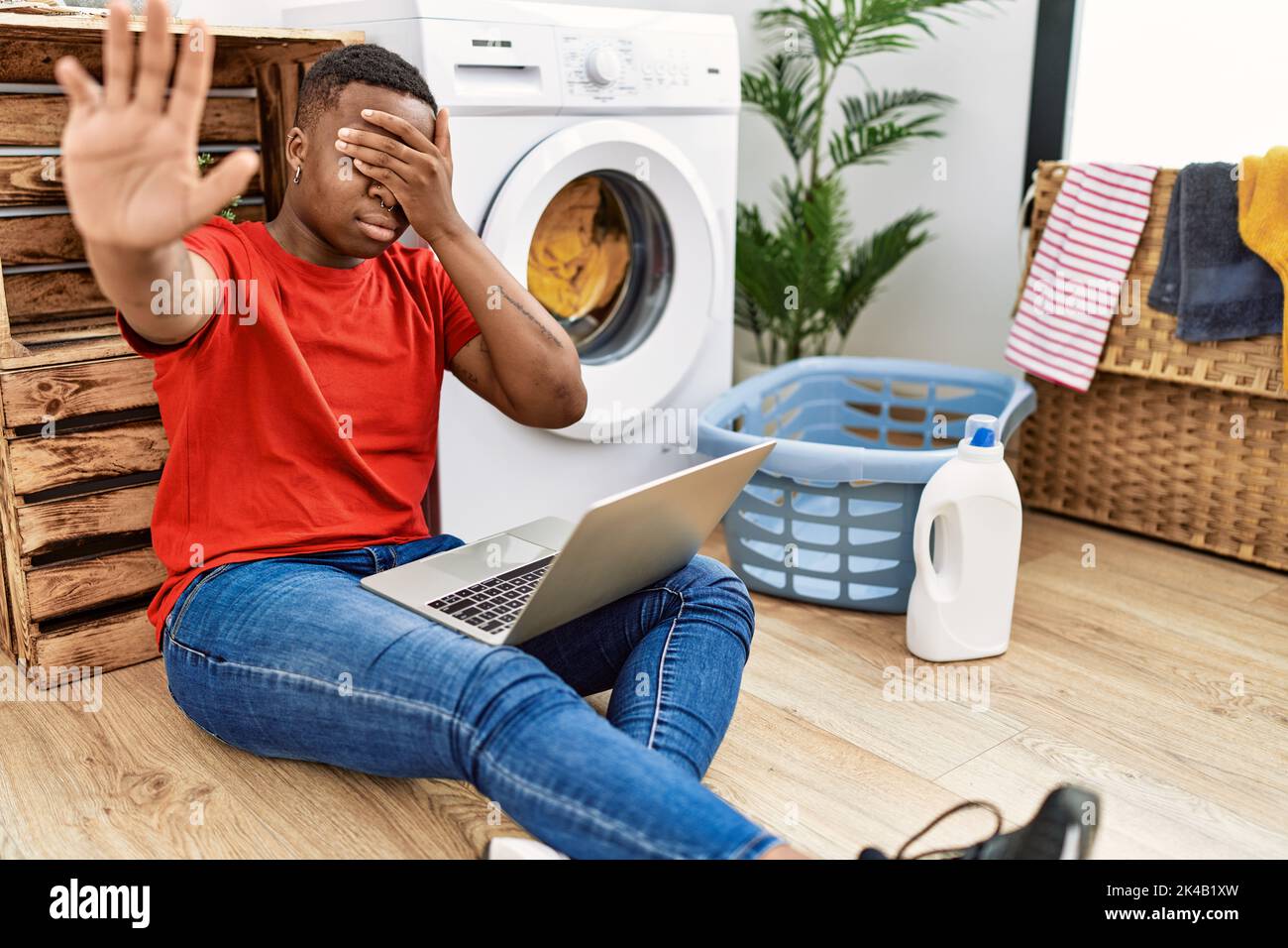 Young african man doing laundry and using computer covering eyes with ...