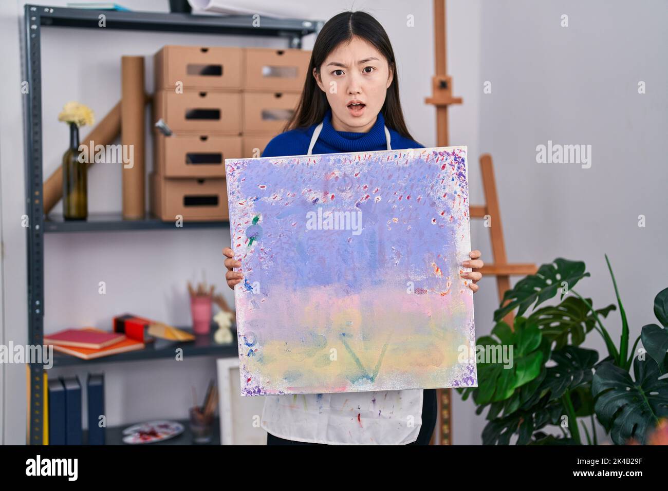 Chinese young woman holding canvas at art studio in shock face, looking ...