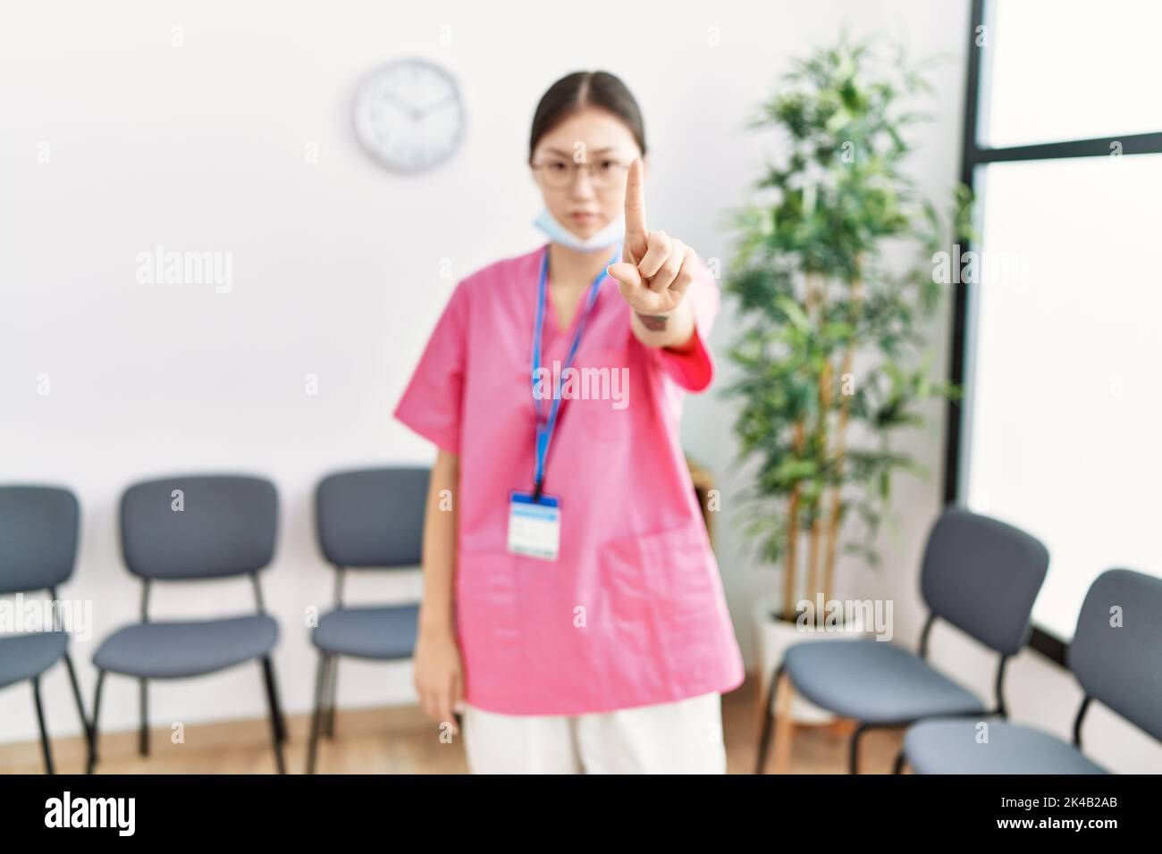 Young asian nurse woman at medical waiting room pointing with finger up ...