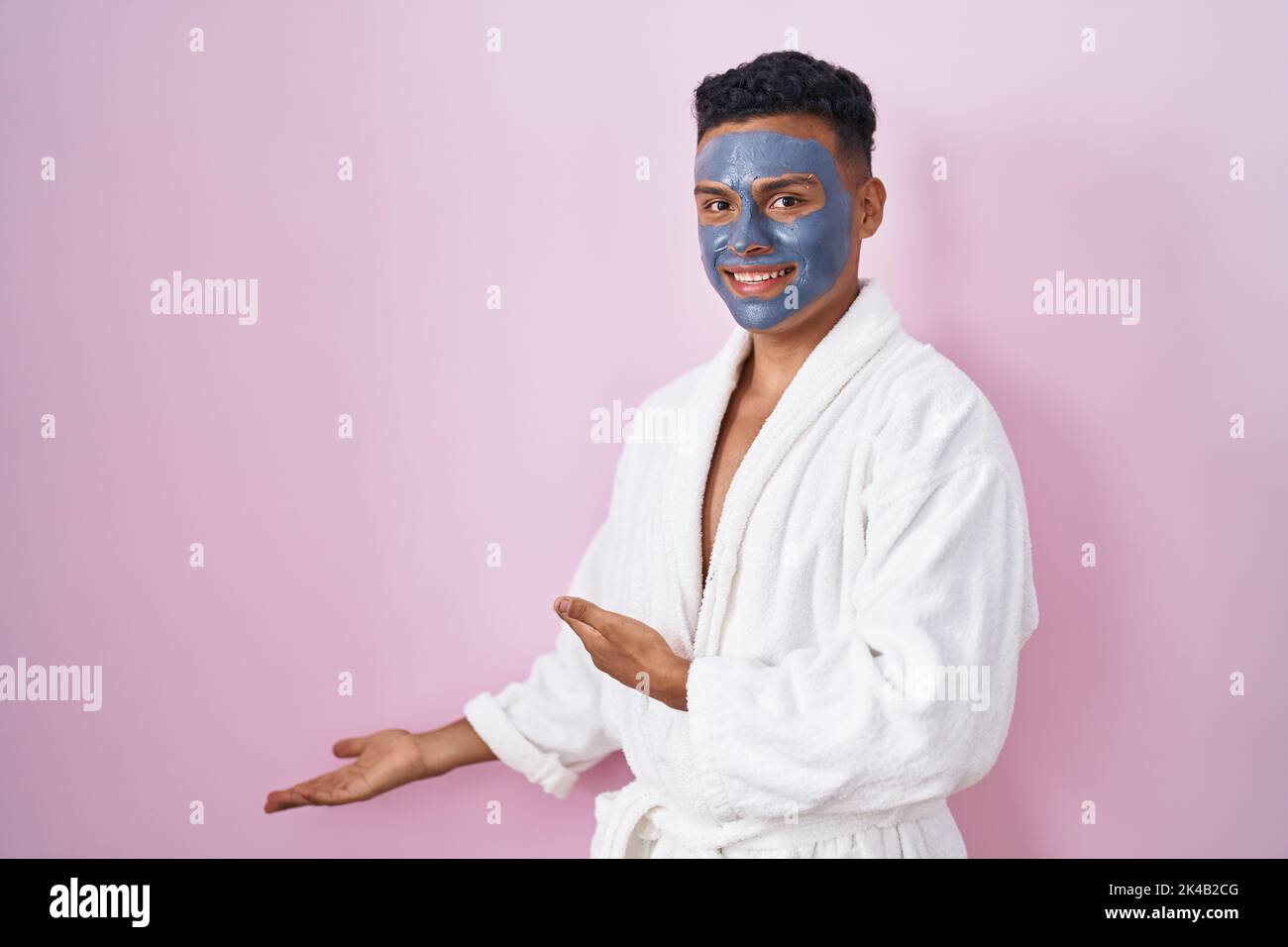 Young hispanic man wearing beauty face mask and bath robe inviting to ...
