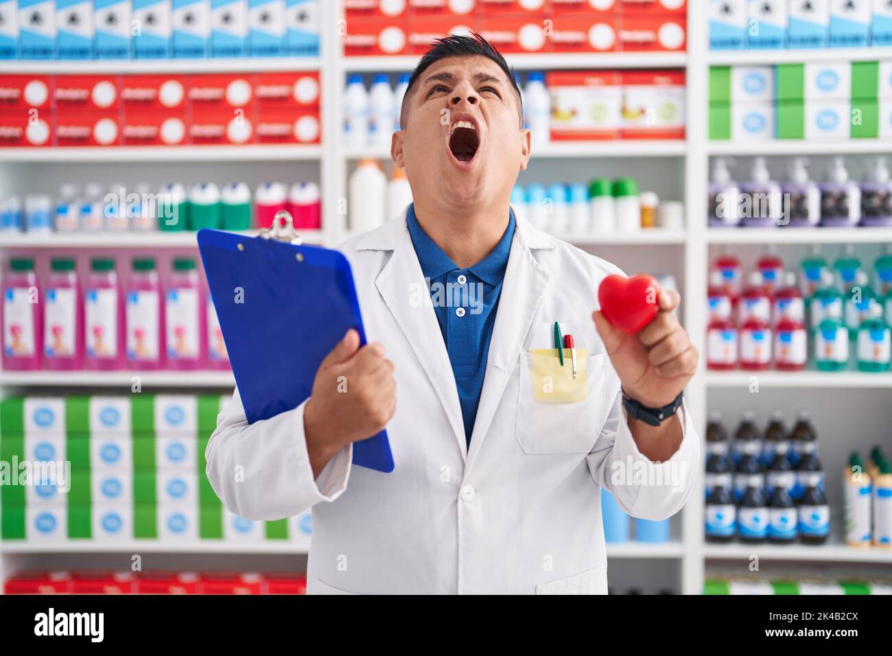 Young hispanic man working at pharmacy drugstore holding heart angry ...