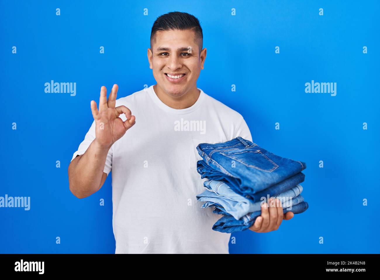 Hispanic young man holding stack of folded jeans doing ok sign with ...