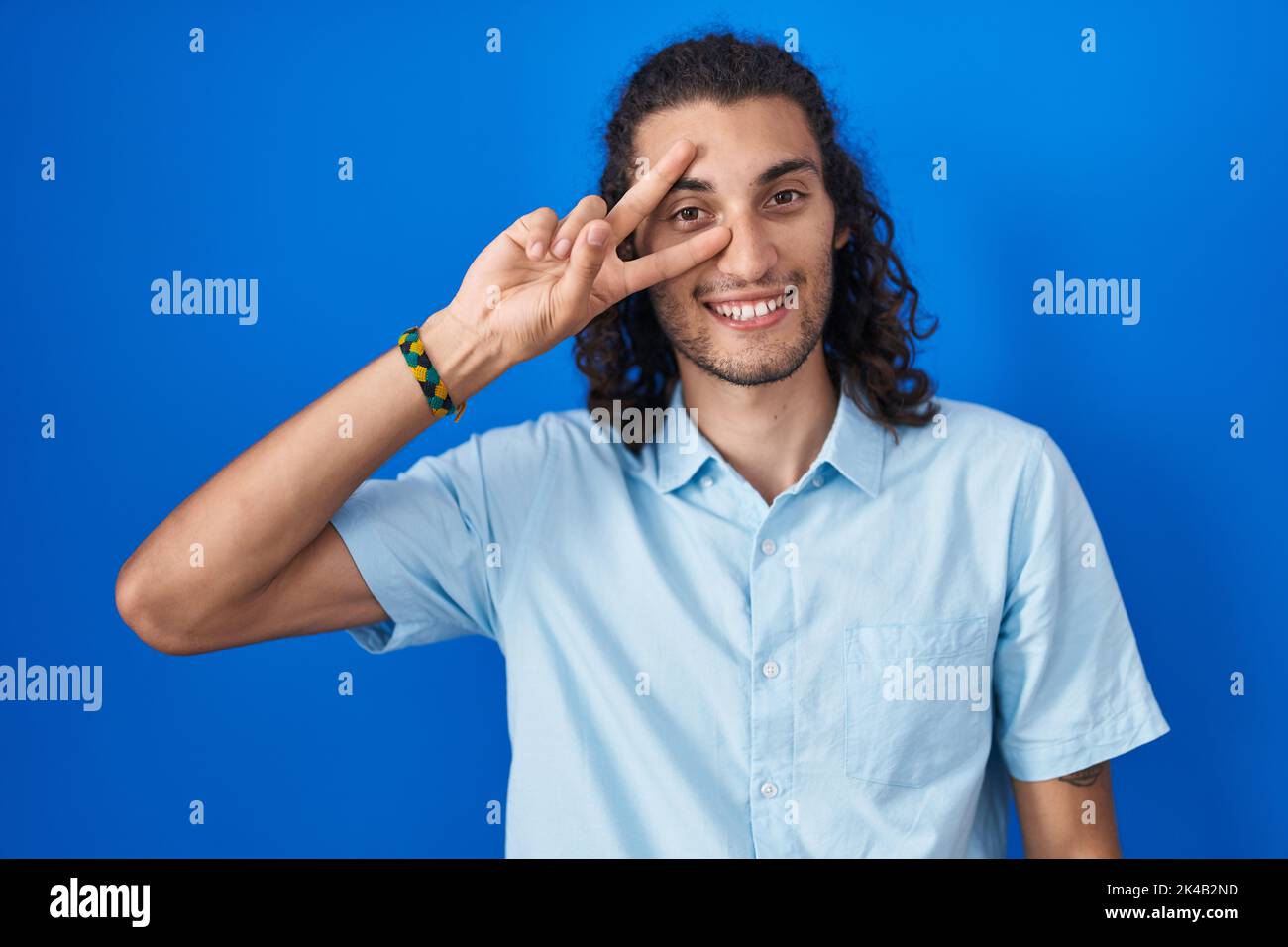 Young hispanic man standing over blue background doing peace symbol ...