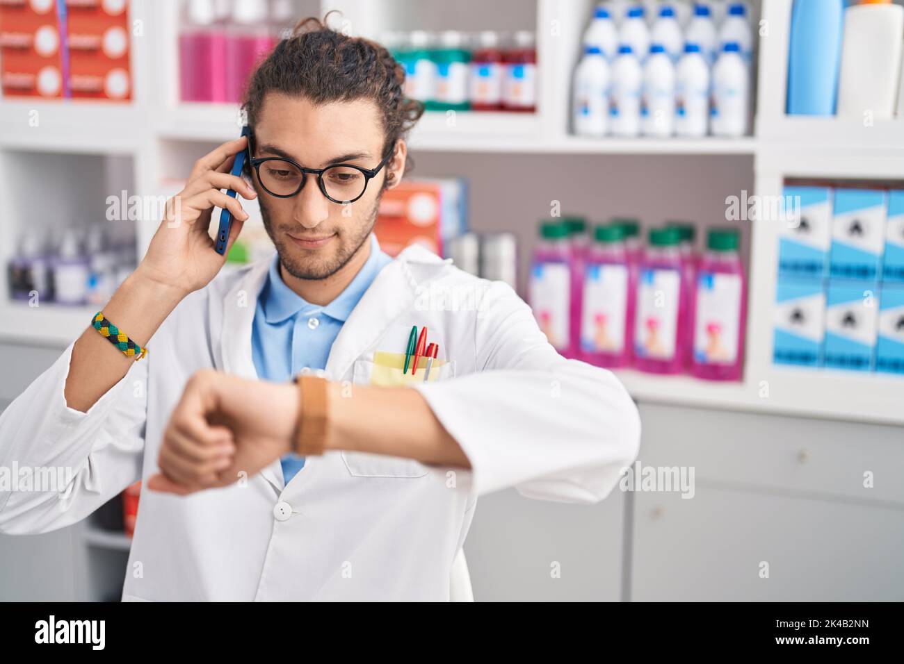 Young hispanic man pharmacist talking on smartphone looking watch at ...