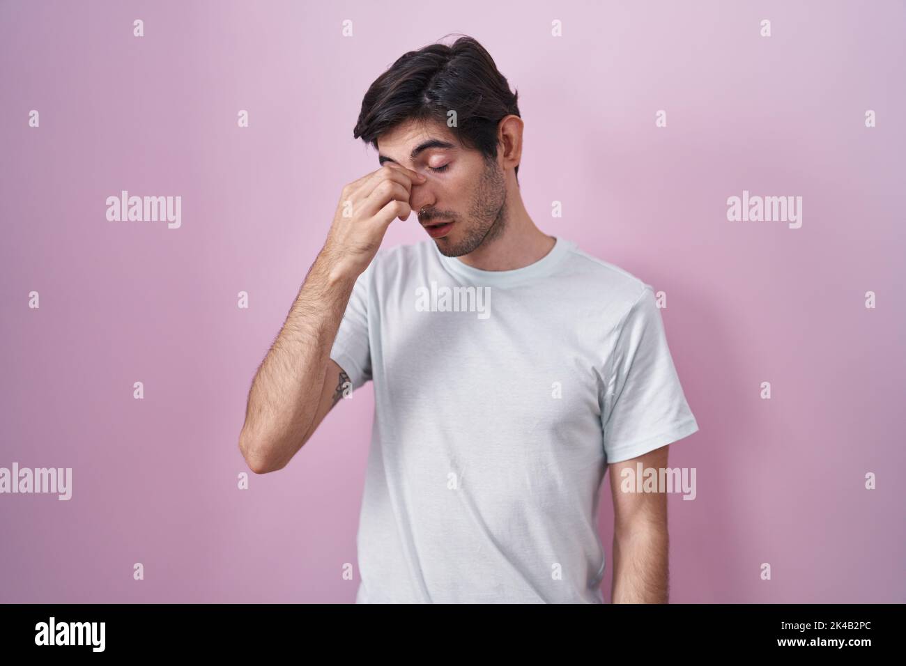 Young hispanic man standing over pink background tired rubbing nose and ...