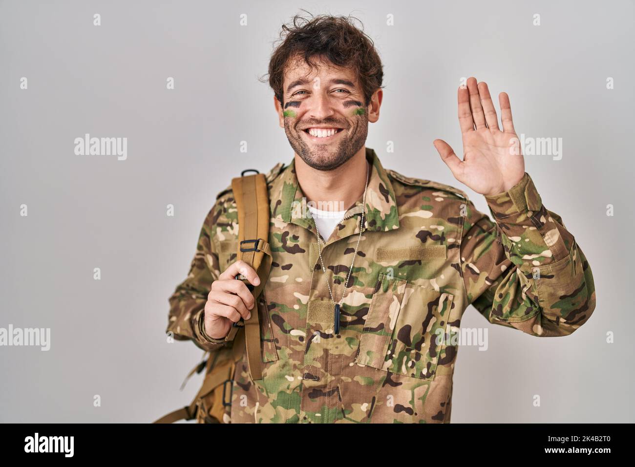 Hispanic young man wearing camouflage army uniform waiving saying hello ...