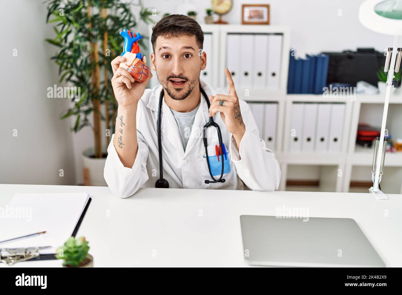 Young doctor holding heart at medical clinic surprised with an idea or ...