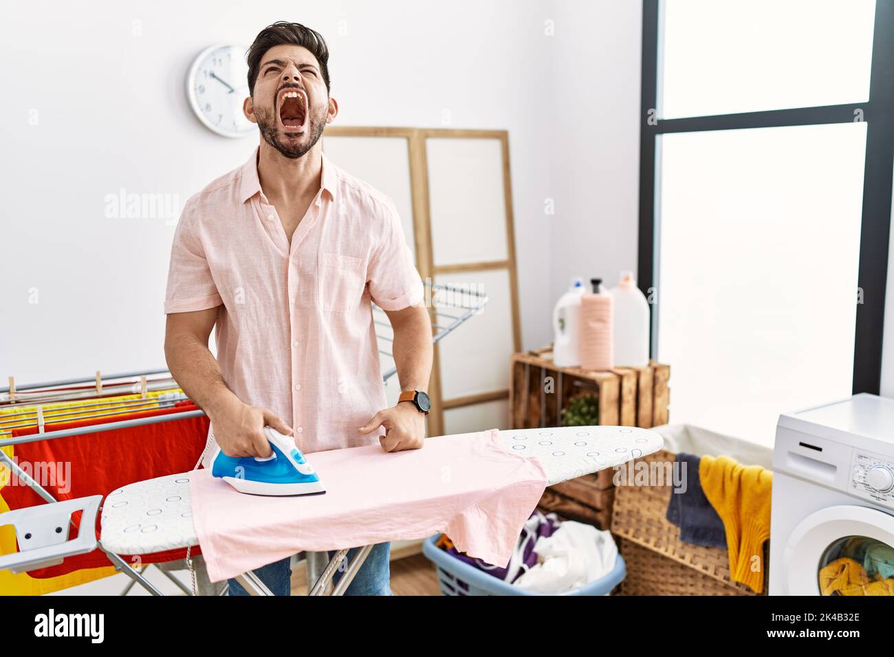 Young man with beard ironing clothes at home angry and mad screaming ...