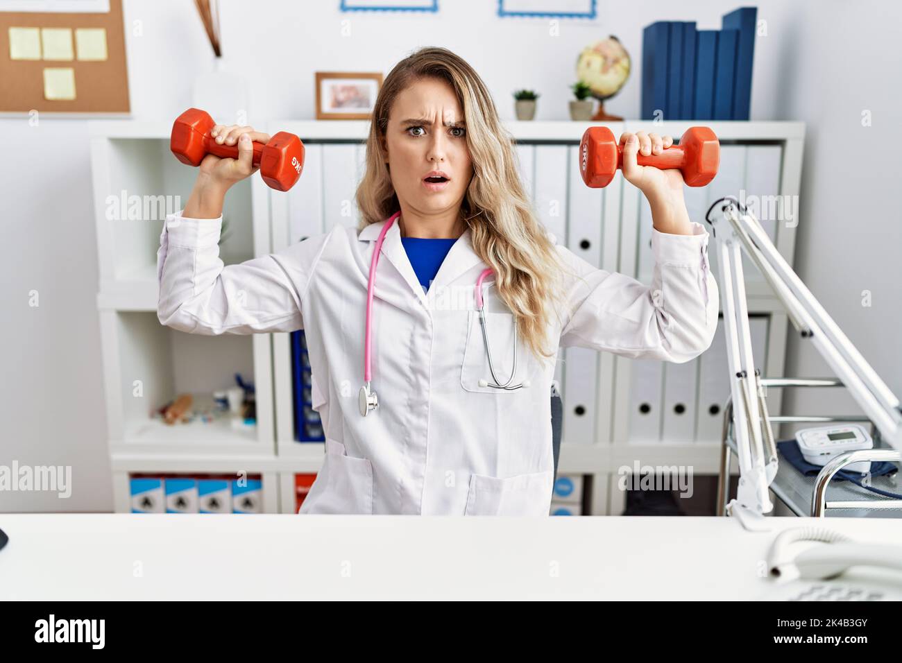 Young beautiful doctor woman holding dumbbells for sport therapy in ...