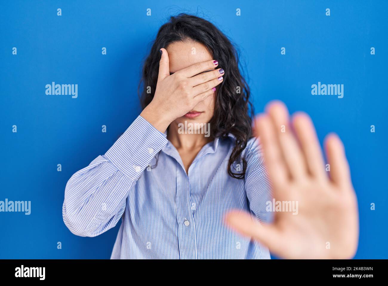 Young brunette woman standing over blue background covering eyes with ...