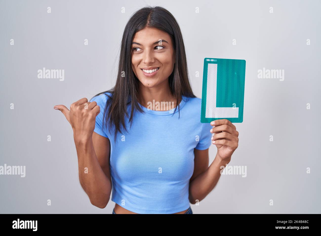 Brunette young woman holding l sign for new driver pointing thumb up to ...