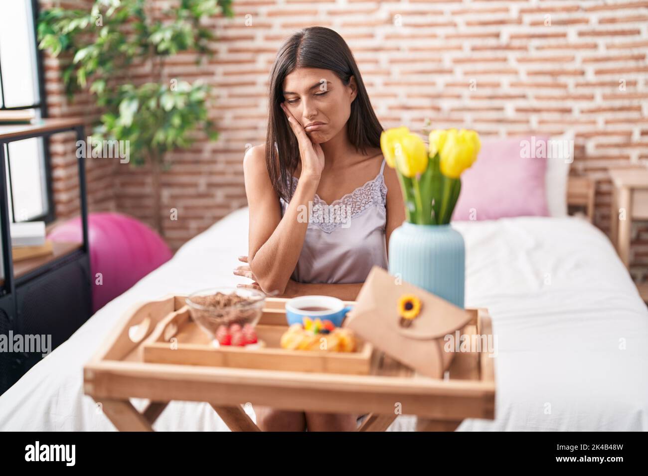 Brunette young woman eating breakfast sitting on the bed thinking looking tired and bored with ...