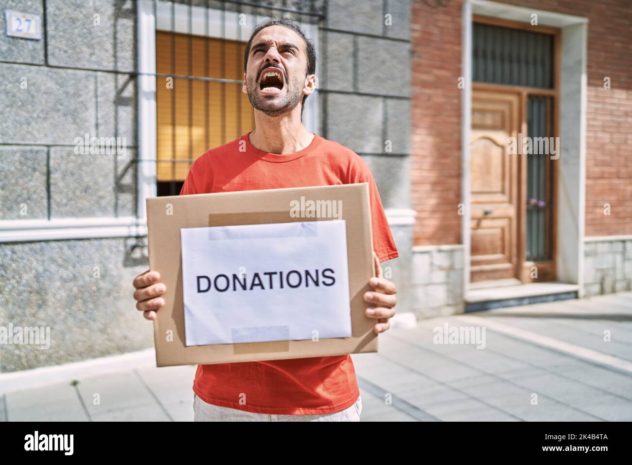 Young hispanic man holding donations box for charity outdoors angry and ...